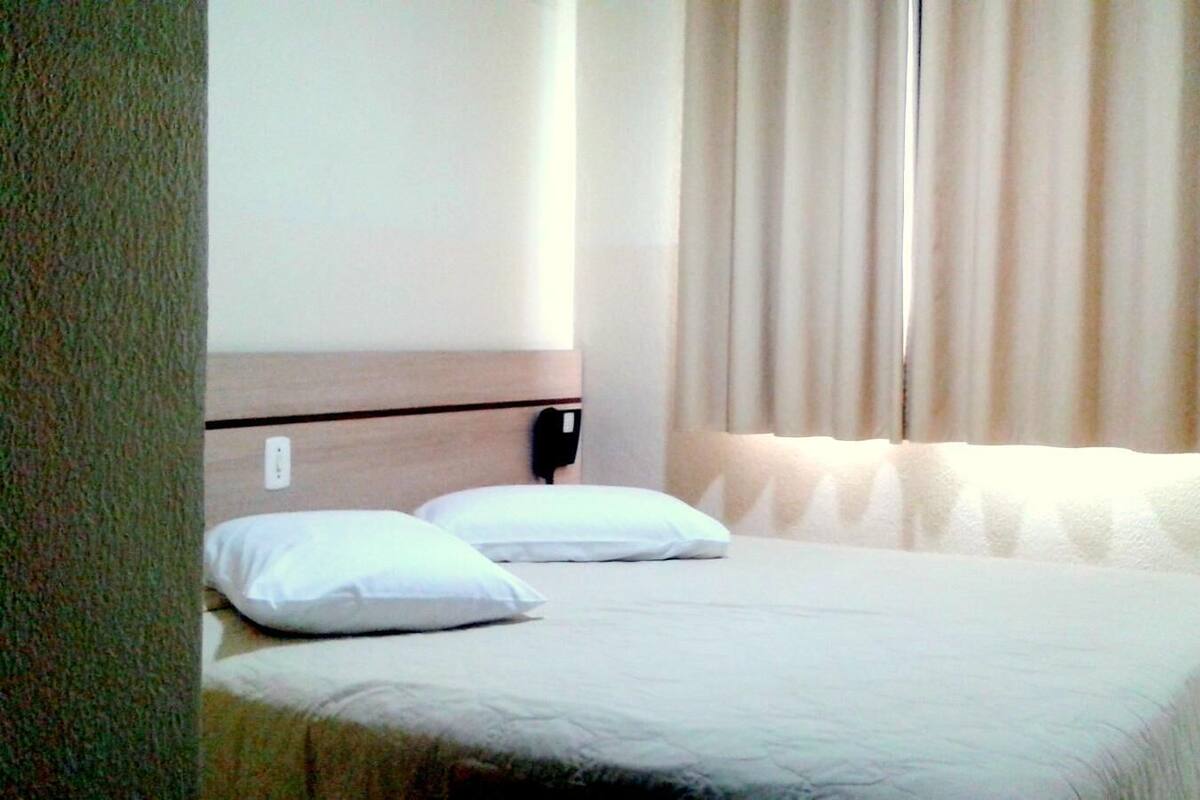 Quarto de hotel com cama, duas almofadas, cabeceira de madeira, mesa de cabeceira e janela com cortinas.