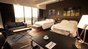 Quarto de hotel com uma cama grande, uma cama menor, um sofá, uma escrivaninha com uma xícara e caneta, e vista para a cidade.