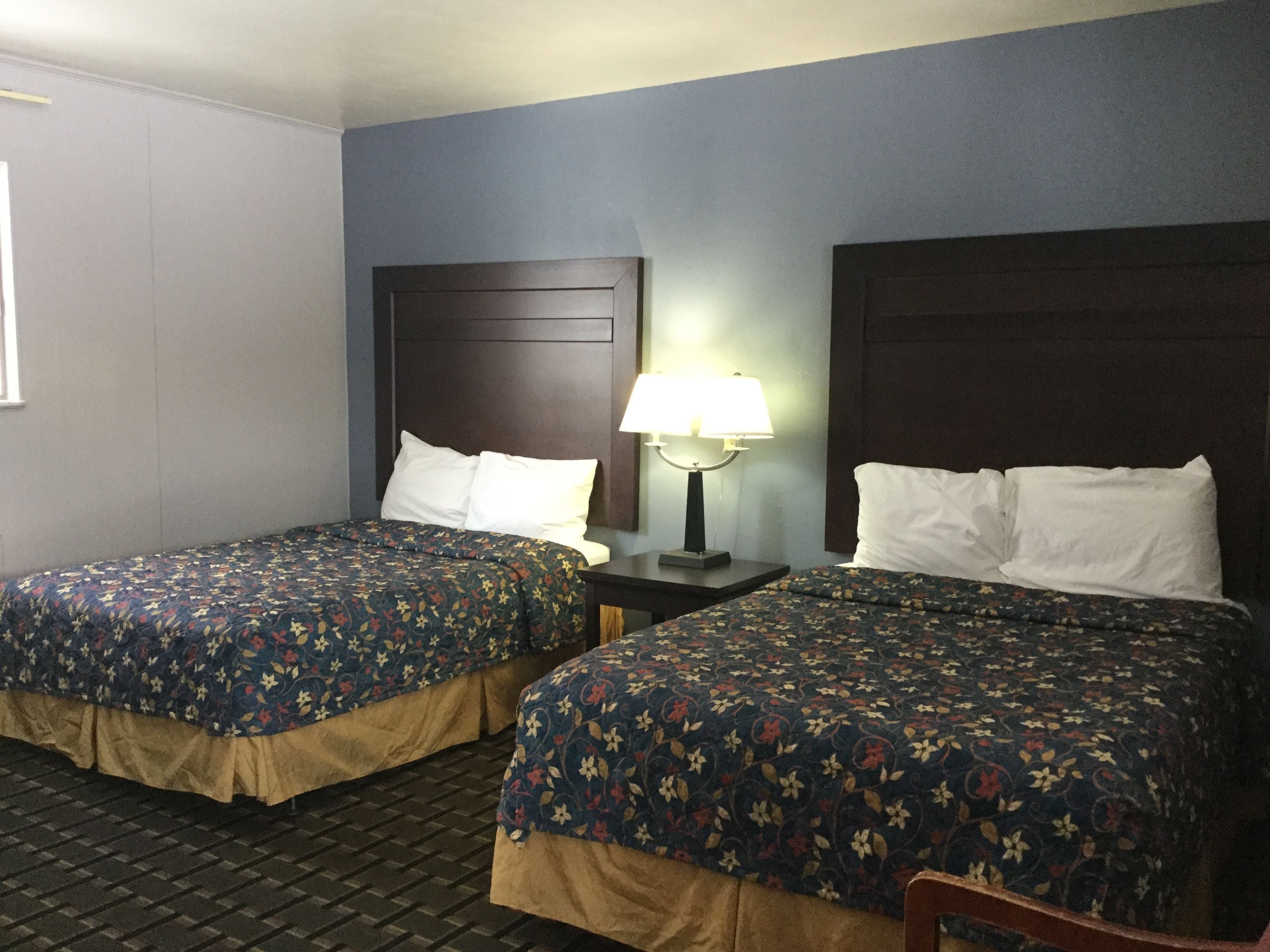 Quarto de hotel com duas camas, cada uma com roupa de cama floral, uma mesinha de cabeceira com abajur e uma janela com persianas.