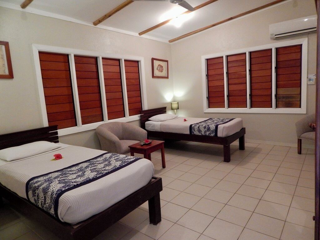 Quarto de hotel com duas camas de solteiro, um pequeno sofá, uma mesa de cabeceira e uma janela com persianas de madeira.