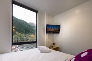 Quarto de hotel com uma janela grande, uma cama com roupa de cama branca, uma pequena mesa de cabeceira com uma planta e um controle remoto, e um quadro na parede.