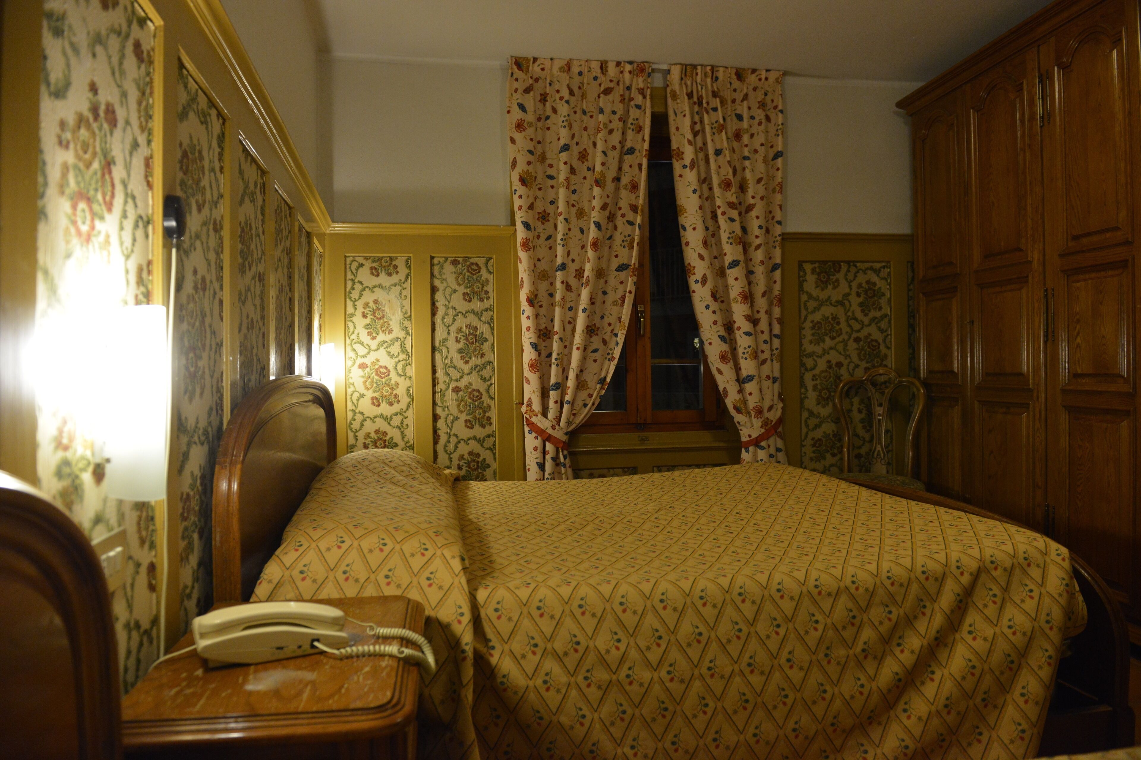 Quarto com cama, guarda-roupa de madeira e mesa de cabeceira com telefone.
