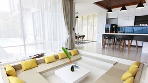 Uma sala moderna com um sofá seccional, uma mesa de centro com um vaso, e uma área de cozinha ao fundo.