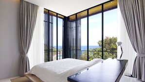Quarto moderno com uma cama grande, mesa de cabeceira e vista para o oceano através de janelas do chão ao teto.