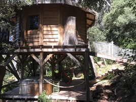 Uma casa na árvore de madeira com uma escada de corda, uma banheira de hidromassagem e uma rede.