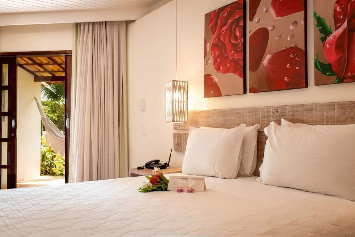 Quarto de hotel com cama, mesa de cabeceira, abajur e uma pintura de rosas.