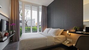 Quarto de hotel moderno com uma cama grande, uma escrivaninha com computador, telefone e uma pequena planta.
