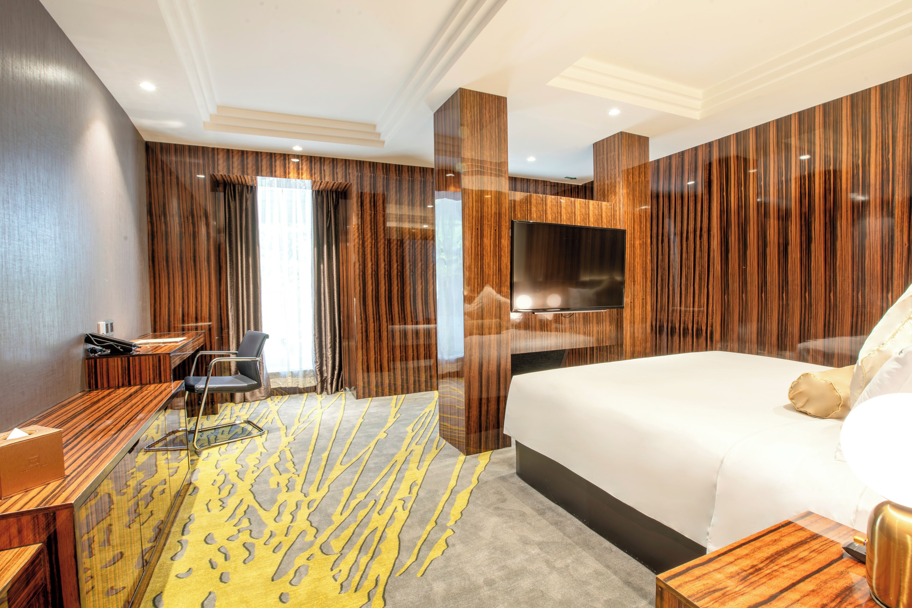 Quarto de hotel com uma cama grande, uma escrivaninha com cadeira, uma televisão de tela plana na parede e um tapete estampado.