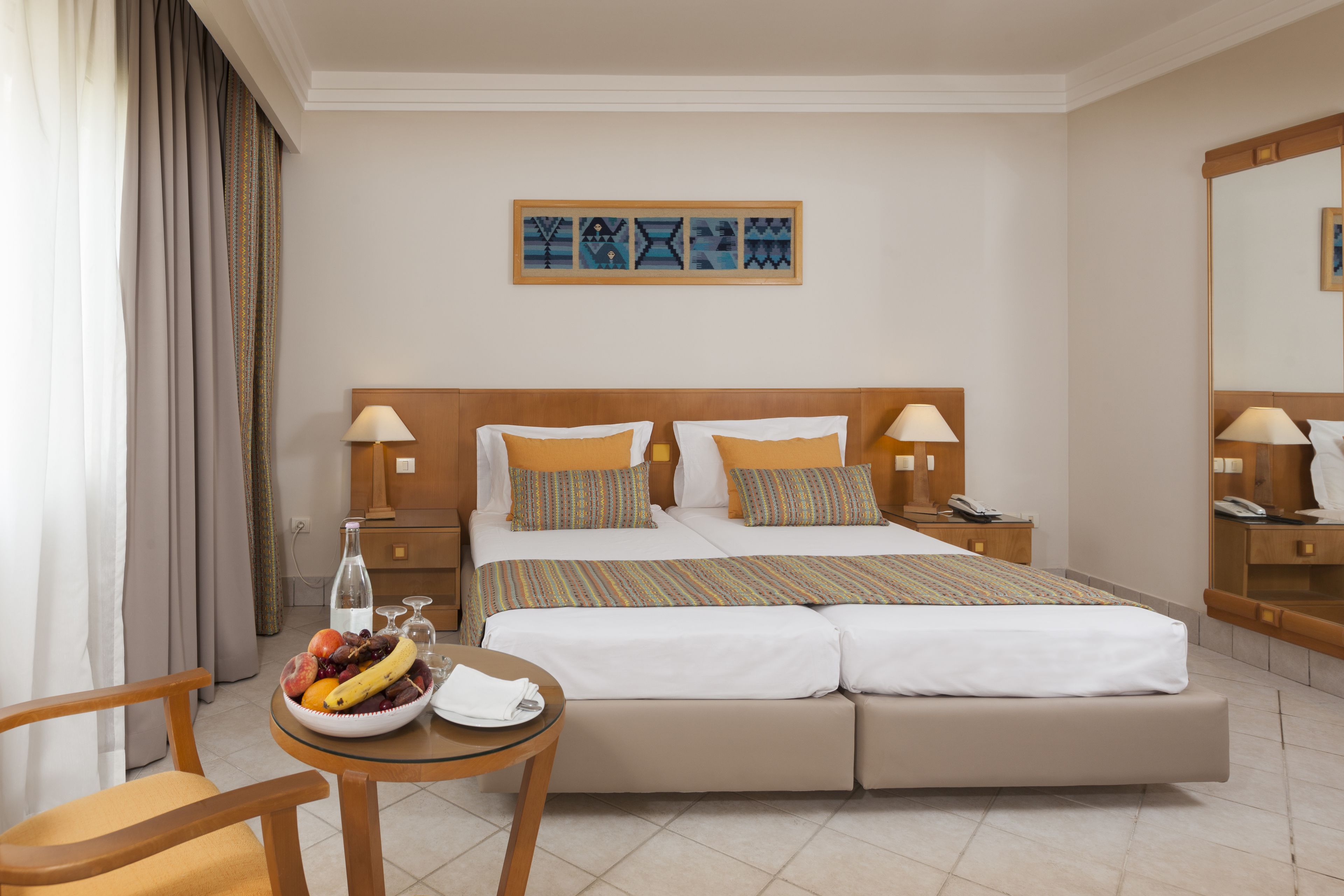 Quarto de hotel com cama de casal, duas mesas de cabeceira com abajures, uma mesinha redonda com uma fruteira e uma garrafa, uma cadeira e um espelho grande.