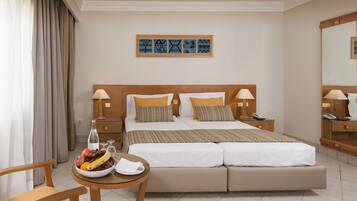 Quarto de hotel com cama de casal, duas mesas de cabeceira com abajures, uma mesinha redonda com uma fruteira e uma garrafa, uma cadeira e um espelho grande.