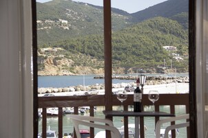 Vista de uma varanda com vista para uma marina, barcos, um litoral rochoso e edifícios numa encosta.