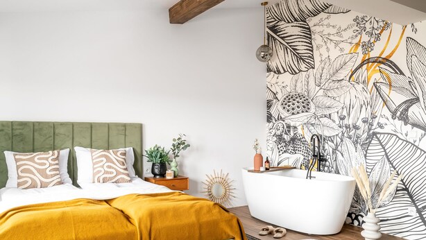 Quarto moderno com cabeceira estofada em verde, cama grande com manta cor mostarda, banheira branca e painel de parede com ilustrações botânicas.