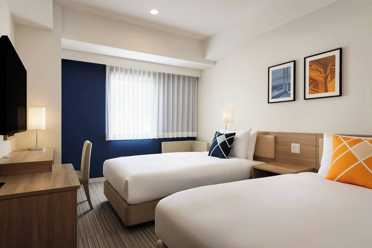 Quarto de hotel com duas camas, uma escrivaninha, uma cadeira, uma televisão e duas imagens emolduradas na parede.