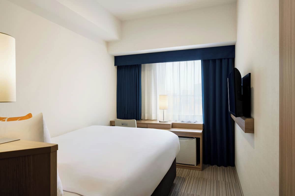 Quarto de hotel com uma cama grande, uma escrivaninha, uma cadeira, TV fixada na parede e janela com cortinas.