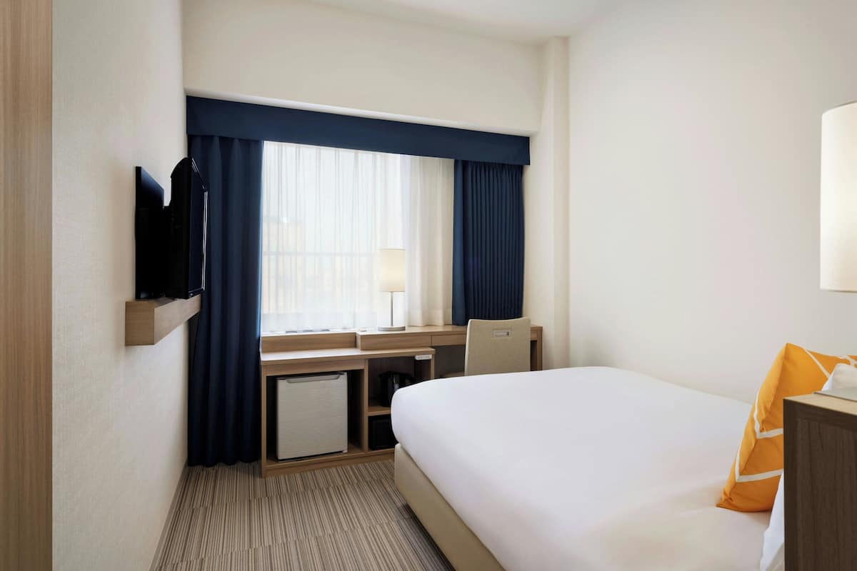 Quarto de hotel com cama, escrivaninha, televisão e janela com cortinas.