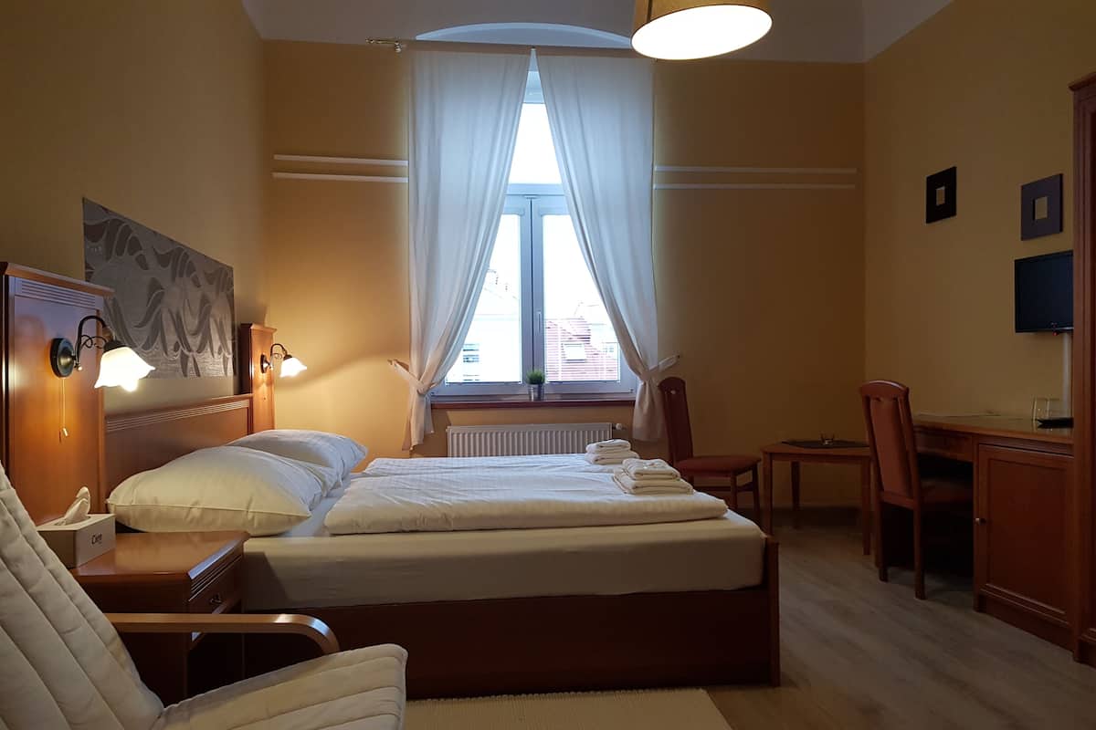 Quarto de hotel com cama, mesa de cabeceira, cadeira, escrivaninha e televisão.