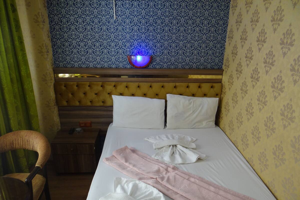 Quarto de hotel com cama, mesa de cabeceira, cadeira e parede com papel de parede estampado.