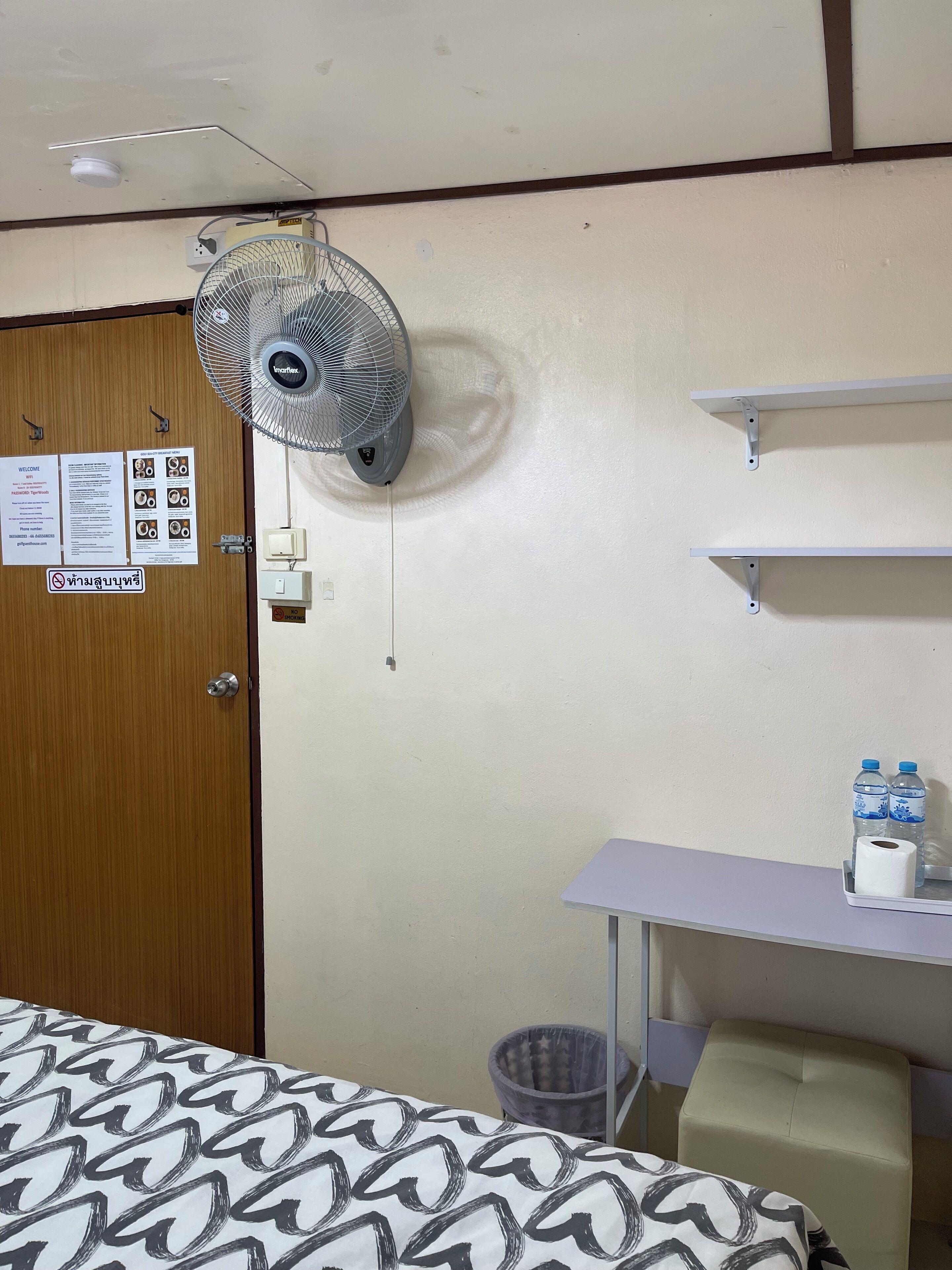 Quarto de hotel com cama, escrivaninha com garrafas de água, lixeira e ventilador fixado na parede.