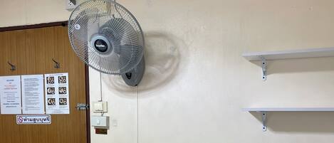 Quarto de hotel com cama, escrivaninha com garrafas de água, lixeira e ventilador fixado na parede.