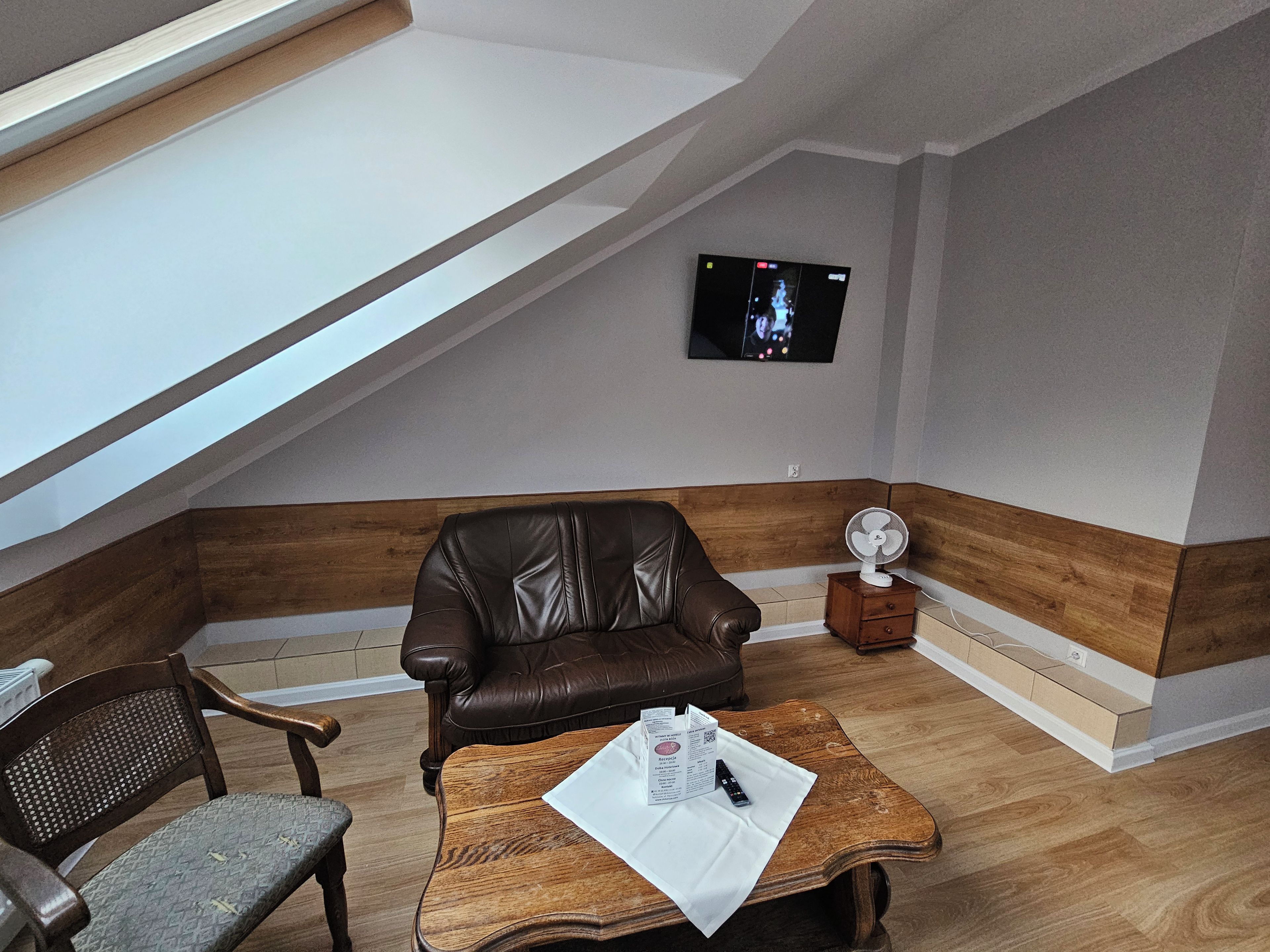 Um quarto com um sofá de couro marrom, uma mesa de centro de madeira, uma televisão fixada na parede e um ventilador sobre uma mesinha de madeira.