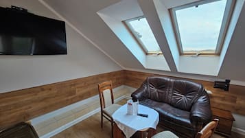 Quarto com sofá de couro marrom, mesa de jantar de madeira com cadeiras, TV de tela plana na parede e janelas do teto que permitem entrada de luz natural.