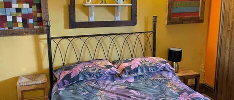 Cama com colcha florida, cabeceira de madeira e estrutura metálica. Há um banquinho pequeno ao lado da cama e uma prateleira com uma planta e um abajur.