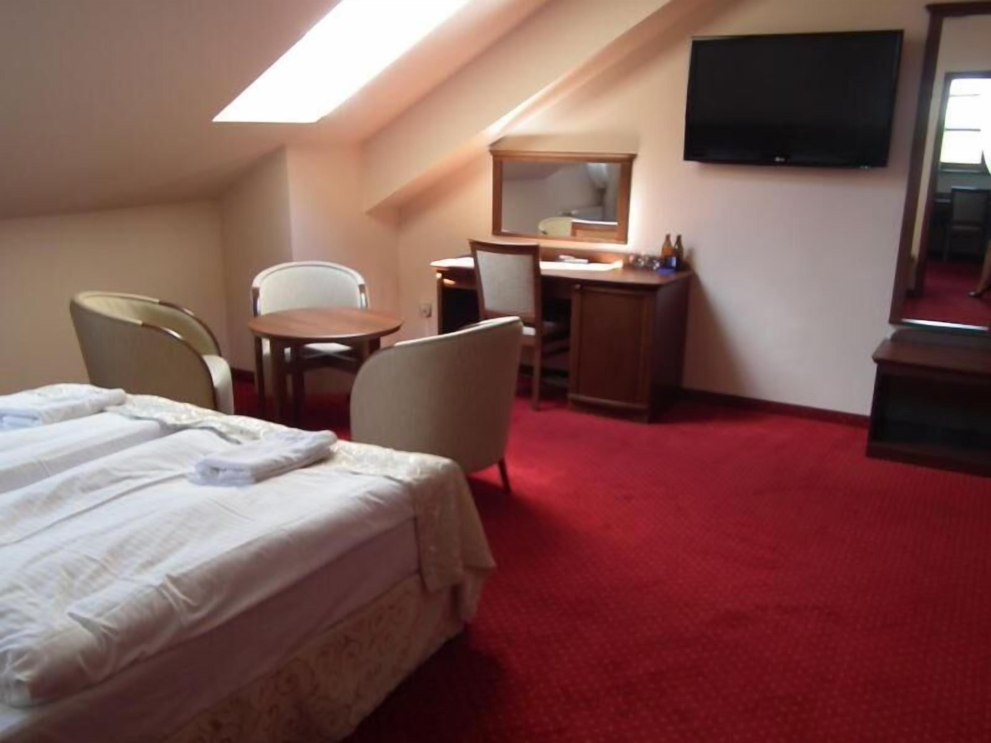 Quarto de hotel com cama, escrivaninha, cadeiras, televisão e espelho.