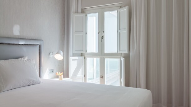 Quarto com cama, mesa de cabeceira, luminária e janela com cortinas.