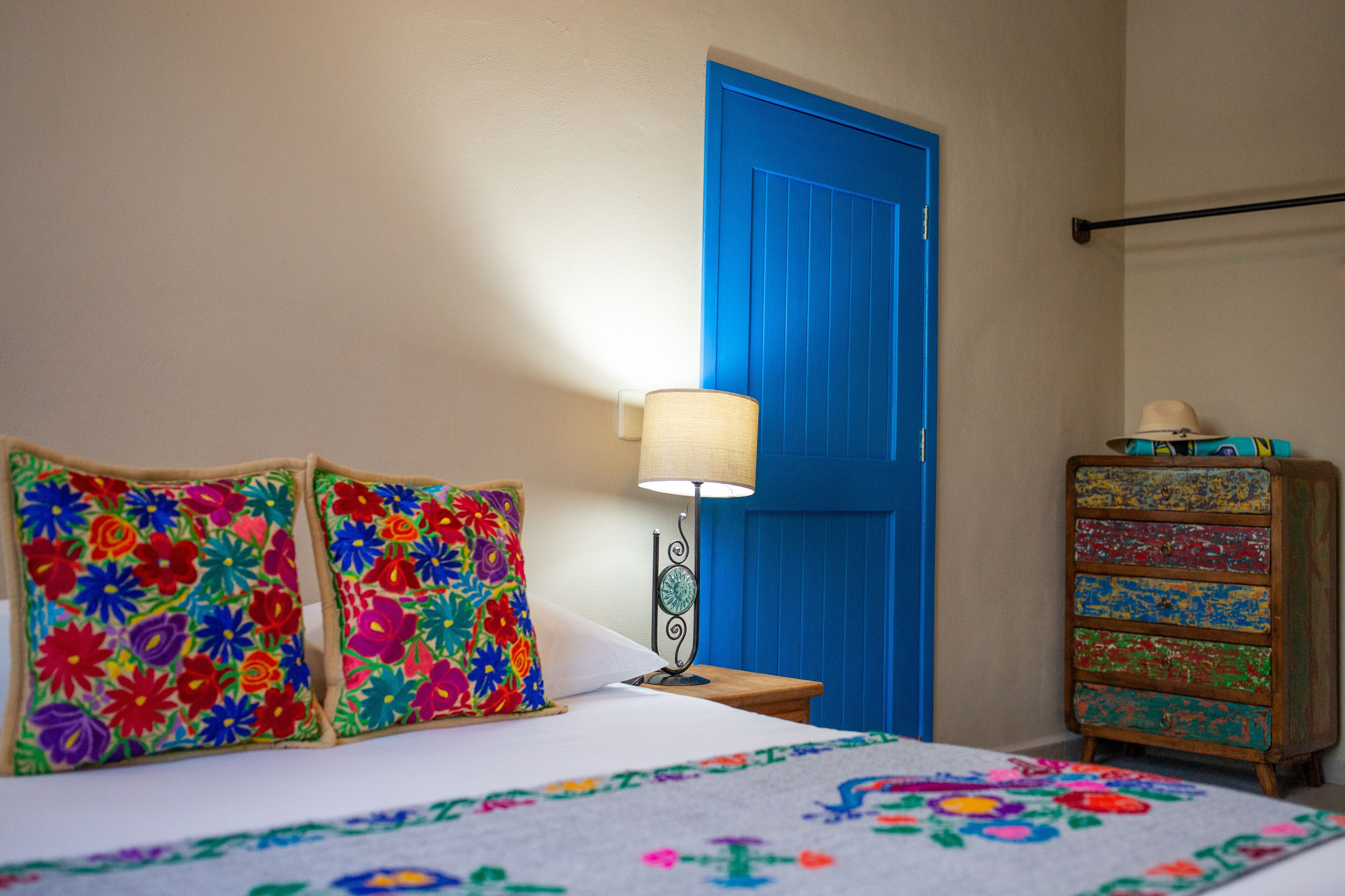 Quarto com cama, almofadas florais coloridas, porta azul, luminária e cômoda de madeira com várias gavetas.