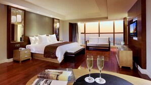Quarto de hotel com uma cama grande, um sofá, uma televisão e uma mesa com duas taças de champanhe.