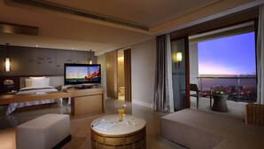 Quarto de hotel moderno com uma televisão grande, varanda com vista para a cidade, área de refeições e um espaço de estar acolhedor.