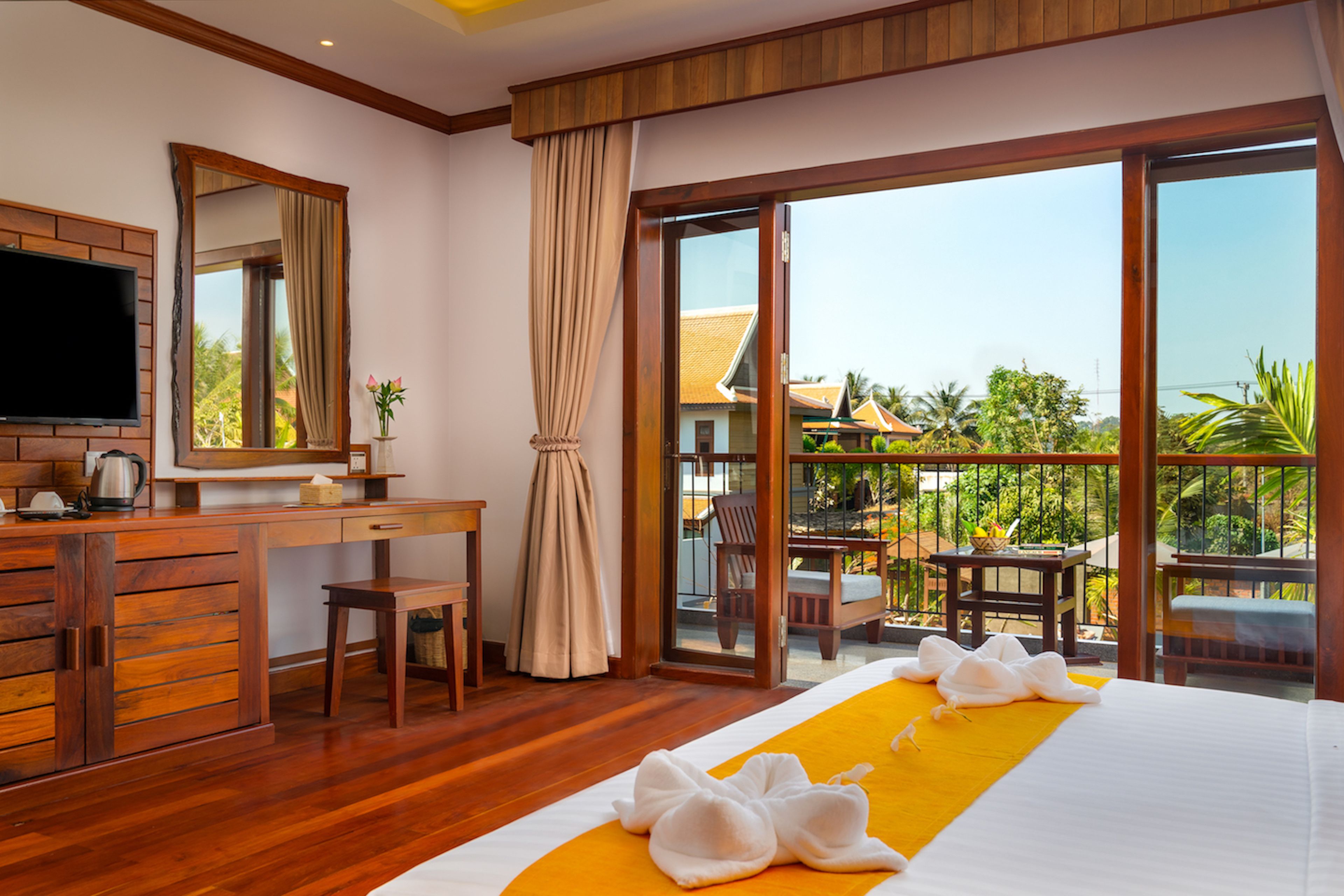 Quarto de hotel com escrivaninha de madeira, televisão, cama com cobre-leito amarelo e varanda com vista para o verde ao redor.