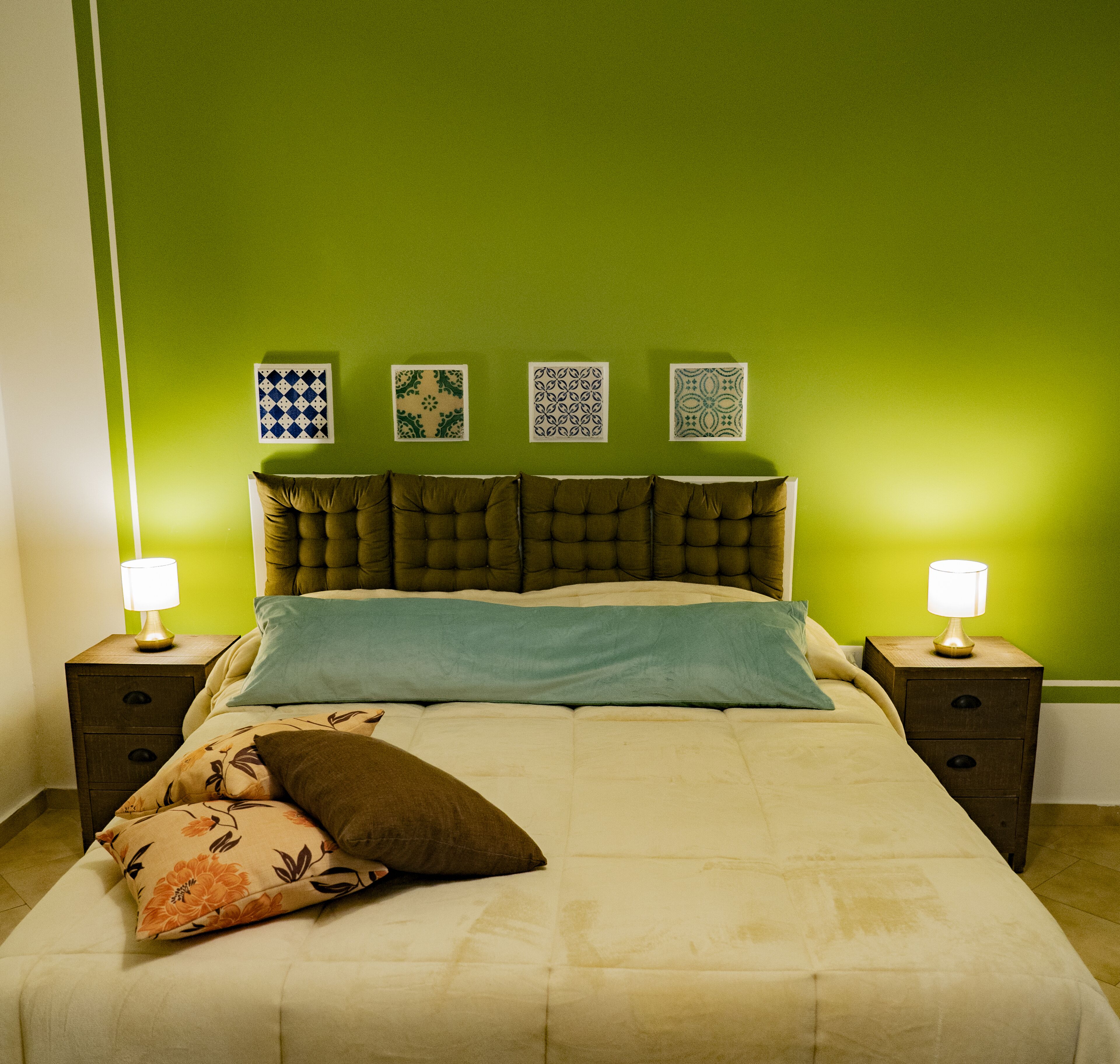 Uma cama bem arrumada com cabeceira verde, duas mesas de cabeceira com abajures e quatro objetos decorativos fixados na parede.