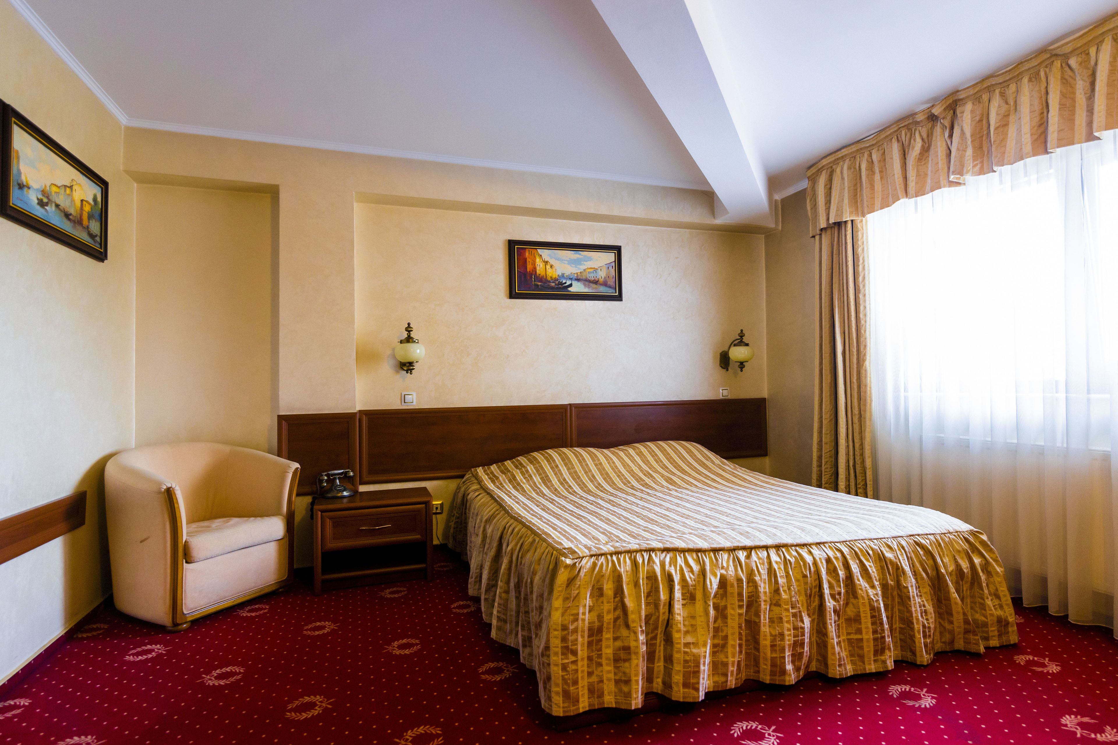 Quarto de hotel com uma cama grande, uma cadeira, uma mesa de cabeceira, um quadro na parede e uma janela com cortinas.