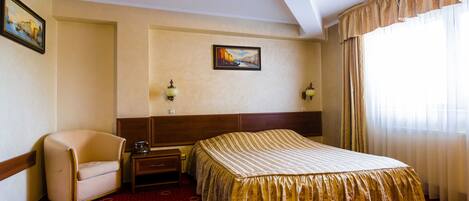 Quarto de hotel com uma cama grande, uma cadeira, uma mesa de cabeceira, um quadro na parede e uma janela com cortinas.