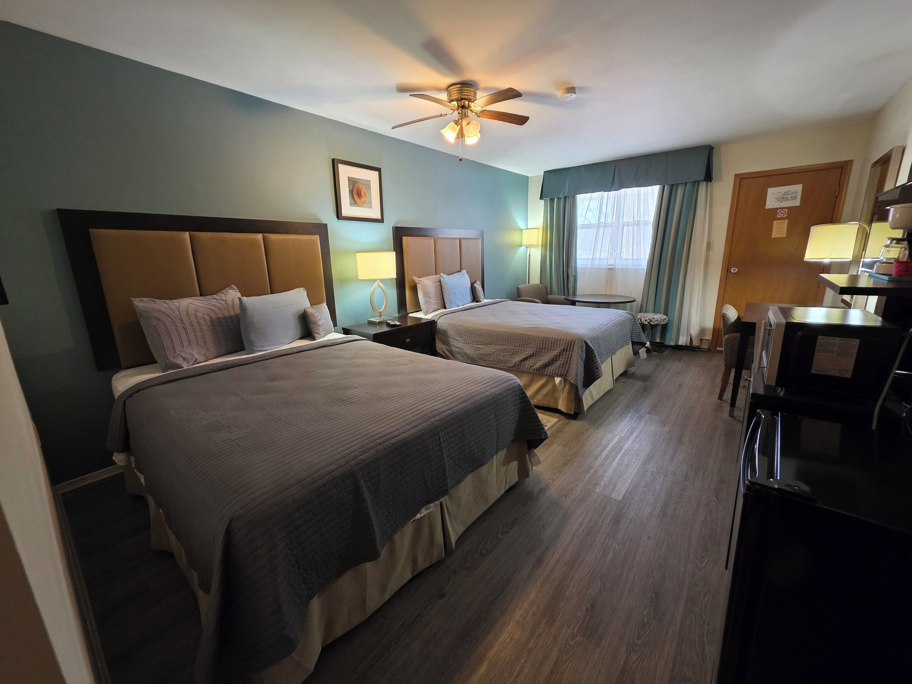 Quarto de hotel com duas camas, um ventilador de teto, uma escrivaninha com cadeira e uma mesinha com abajur.