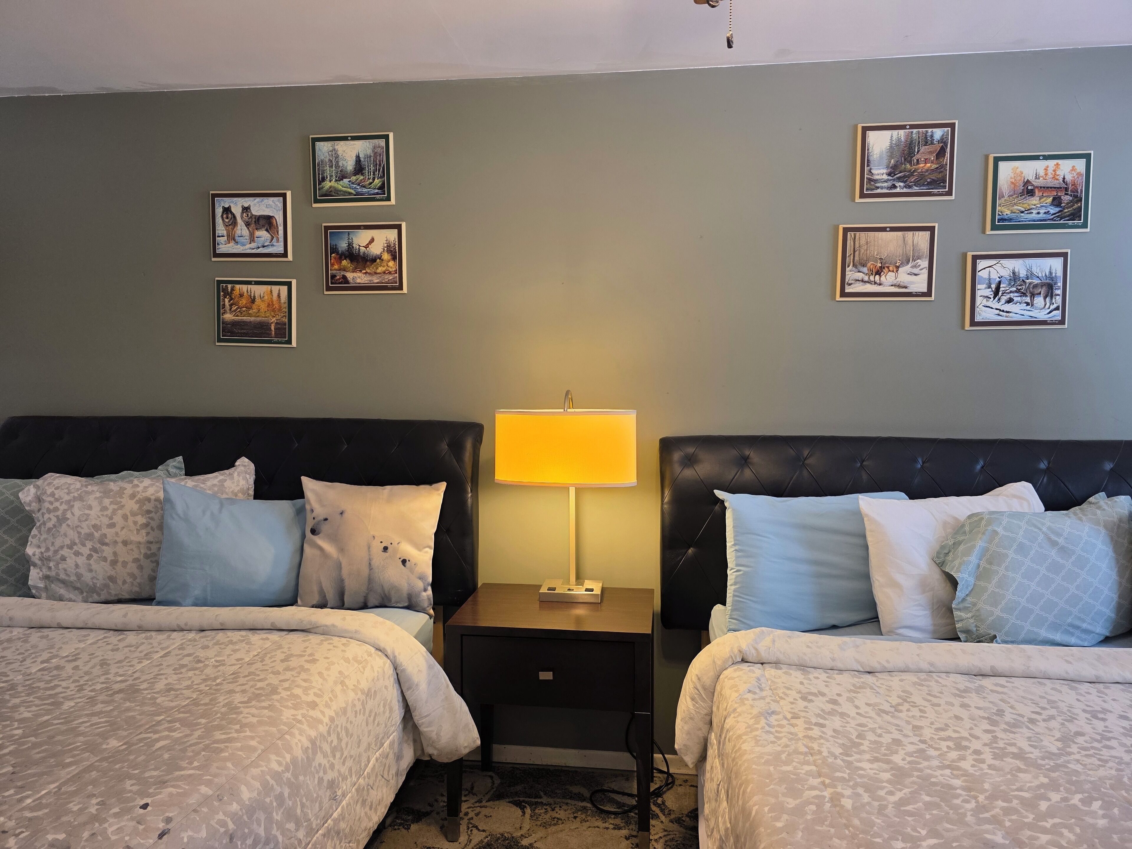 Quarto de hotel com duas camas, uma mesa de cabeceira com abajur e várias fotos emolduradas na parede.