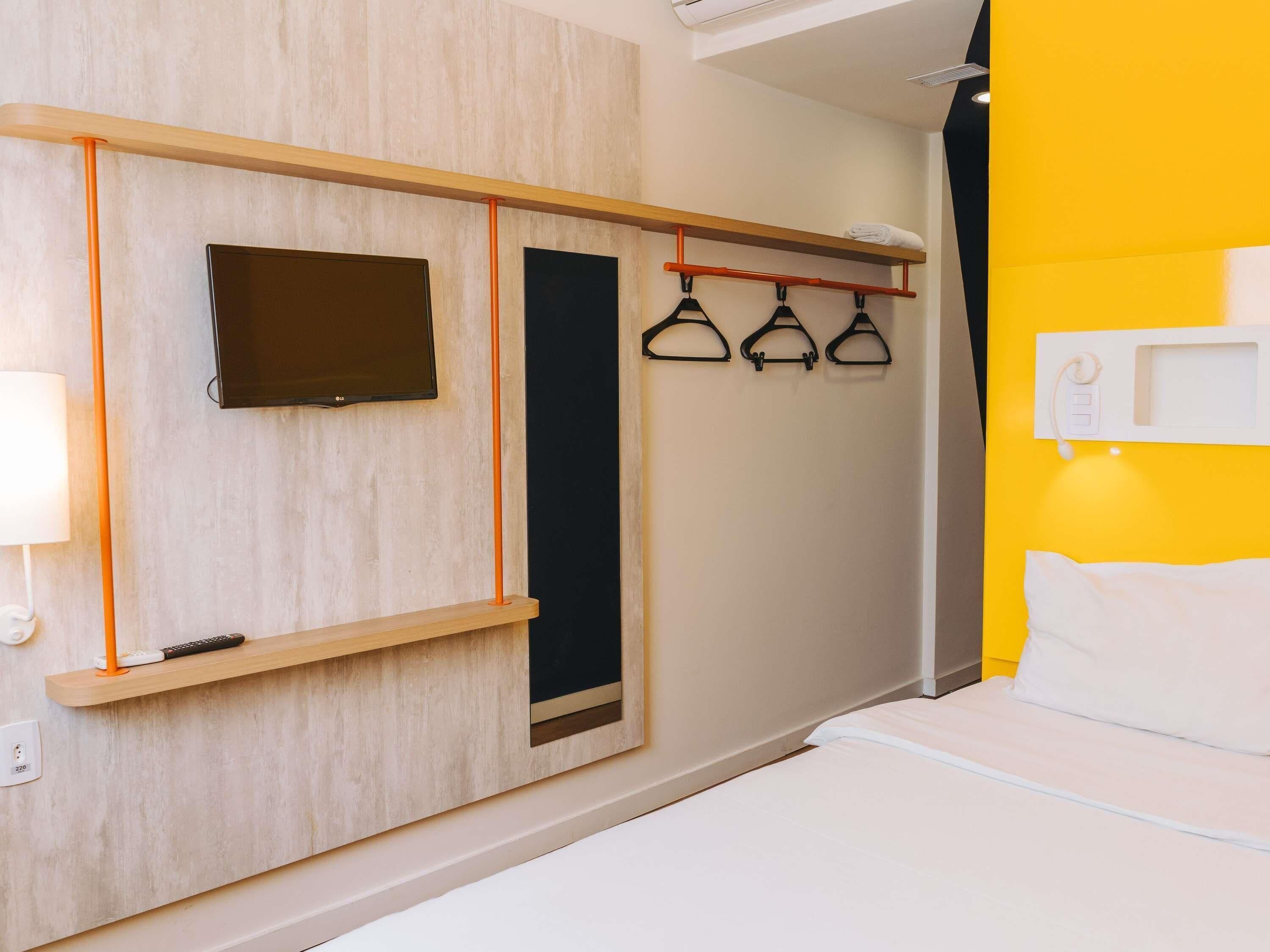 Quarto de hotel com cama, televisão fixada em um painel de madeira, cabideiro com ganchos e micro-ondas na parede.