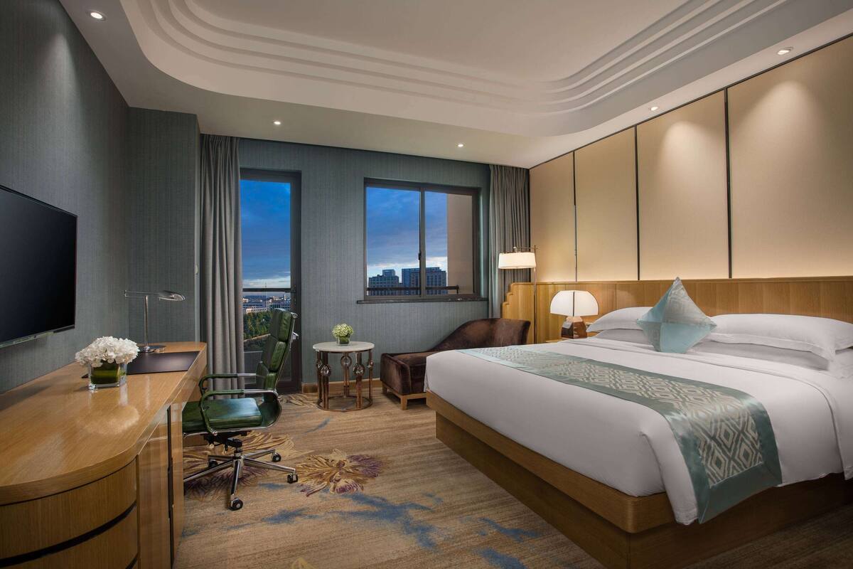 Quarto de hotel com uma cama grande, uma escrivaninha com cadeira, televisão e vista para a cidade.