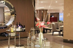 Um saguão de hotel com um espelho redondo, uma mesa de vidro com um vaso de flores e uma área de bar com garrafas.