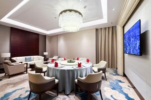 Uma sala de conferências de hotel com uma mesa redonda, cadeiras, um lustre, uma televisão na parede e um tapete estampado.