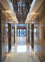 Um lobby de hotel moderno, com piso de mármore polido, entrada central com portas de vidro e luminária pendente.