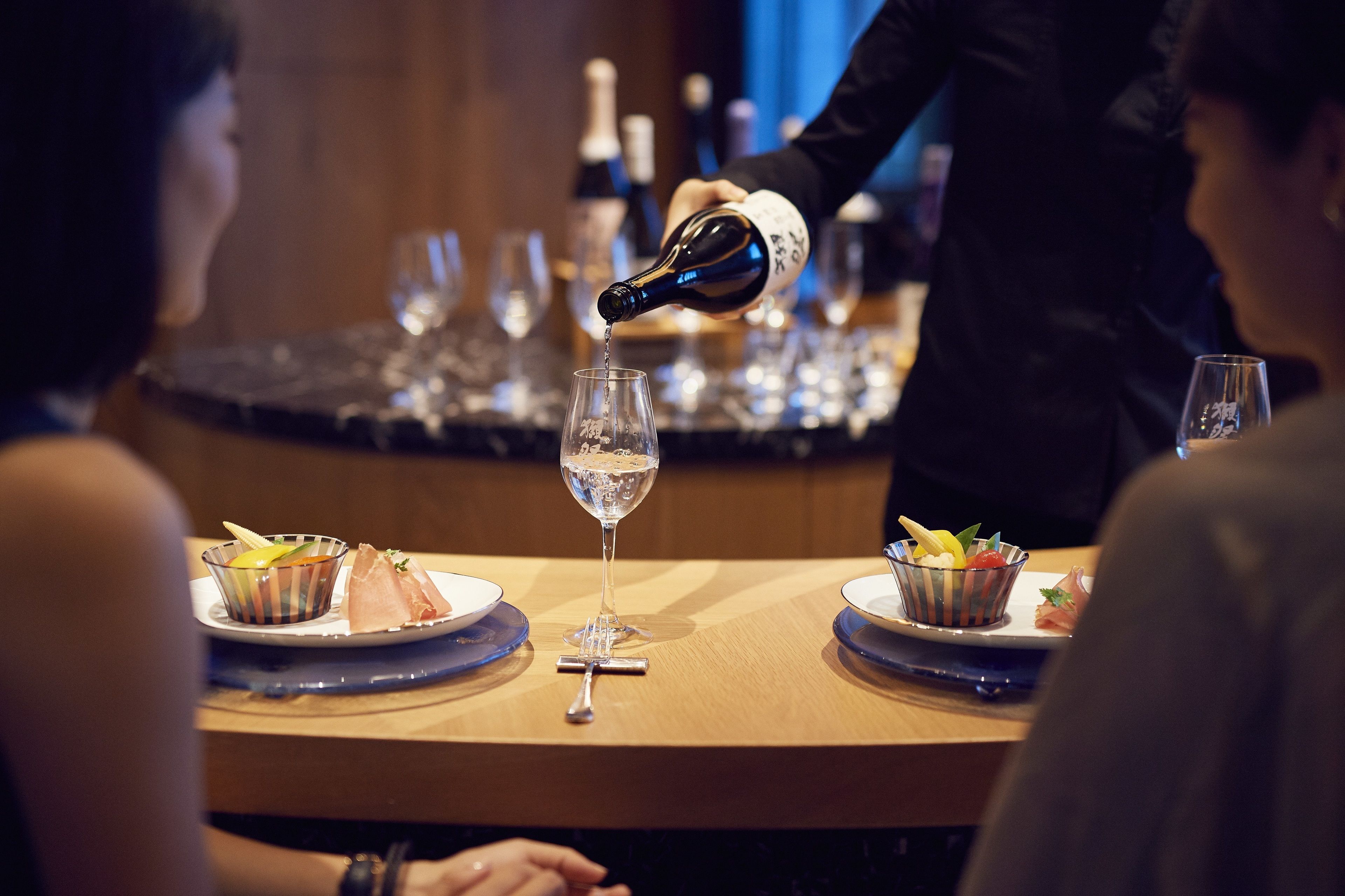 Um bar com alguém servindo uma bebida em um copo, dois pratos com comida e um balcão de bar cheio de copos e garrafas.