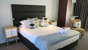 Cama bem arrumada com cabeceira escura, roupa de cama branca e almofadas estampadas. Há uma pequena mesa de cabeceira com luminária, um criado-mudo com gavetas e uma cadeira com almofada.