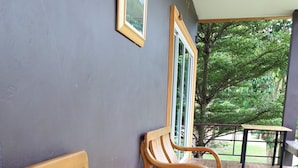 Uma área de estar coberta ao ar livre com bancos de madeira e uma mesa, emoldurada por uma janela com vista para o verde.
