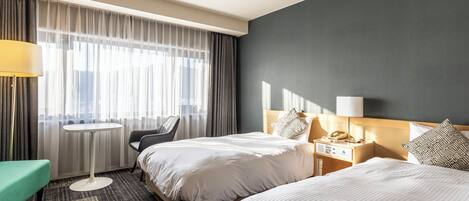 Quarto de hotel com duas camas, uma mesa pequena, uma cadeira e vista para a cidade através de cortinas translúcidas.