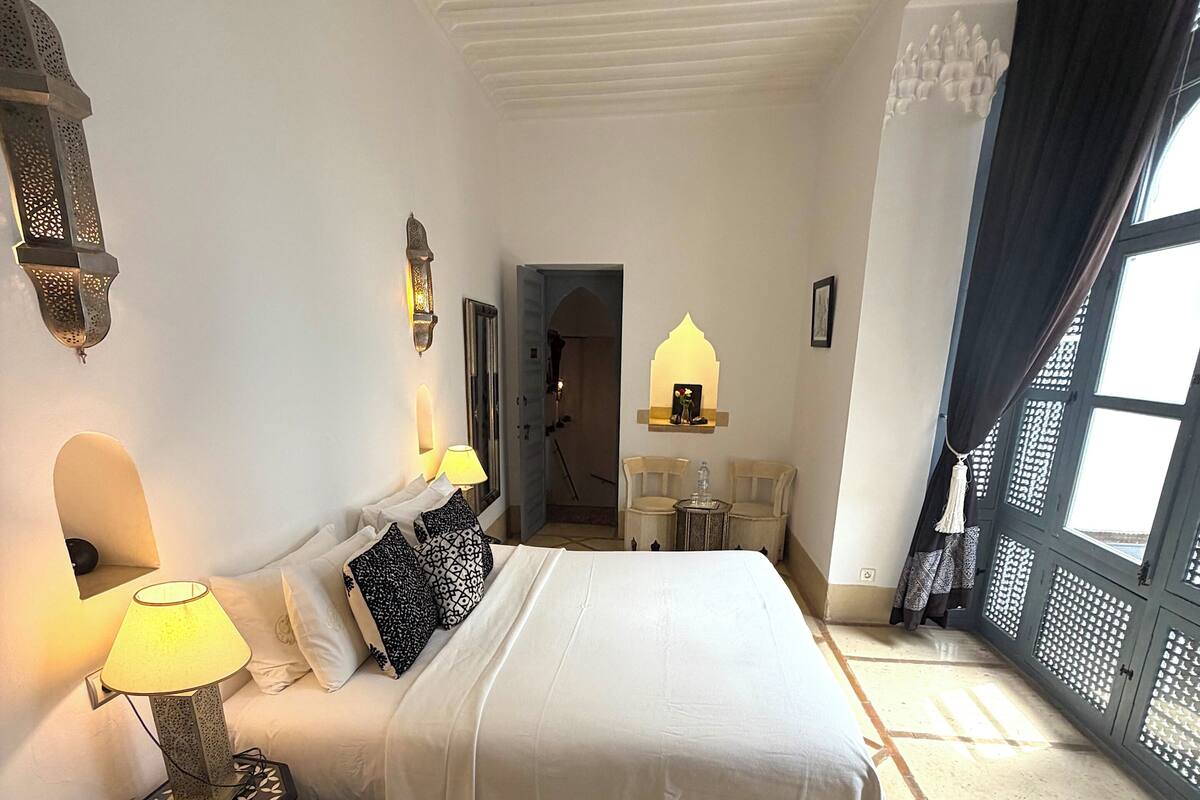 Quarto com cama, duas mesas de cabeceira com abajures, um tapete, uma prateleira na parede com um quadro e uma janela com cortinas.