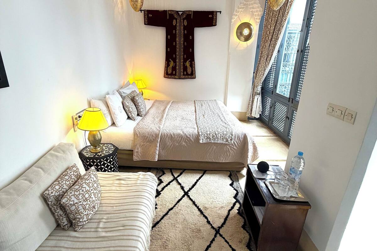 Um quarto com cama, mesas de cabeceira, um sofá, um tapete estampado, luminárias pendentes e um espelho decorativo.