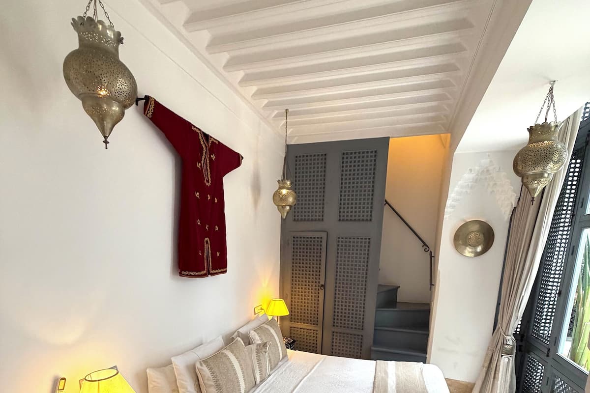 Um quarto com uma cama, duas luminárias na parede, uma peça de roupa vermelha, um tapete estampado e uma escada que leva a outro cômodo.