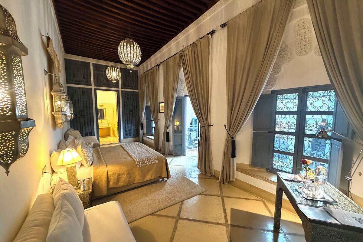 Quarto com cama, luminárias de cabeceira, um sofá, uma mesa com um vaso e vista pela janela.